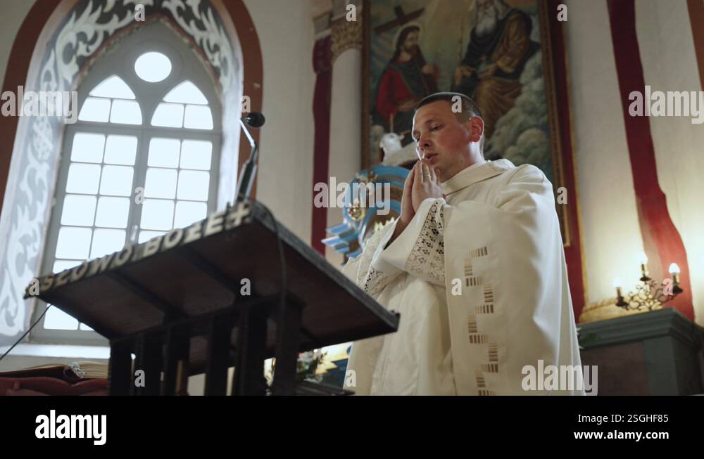 Priest in white attire folded palms on chest and prays behind pulpit in ...