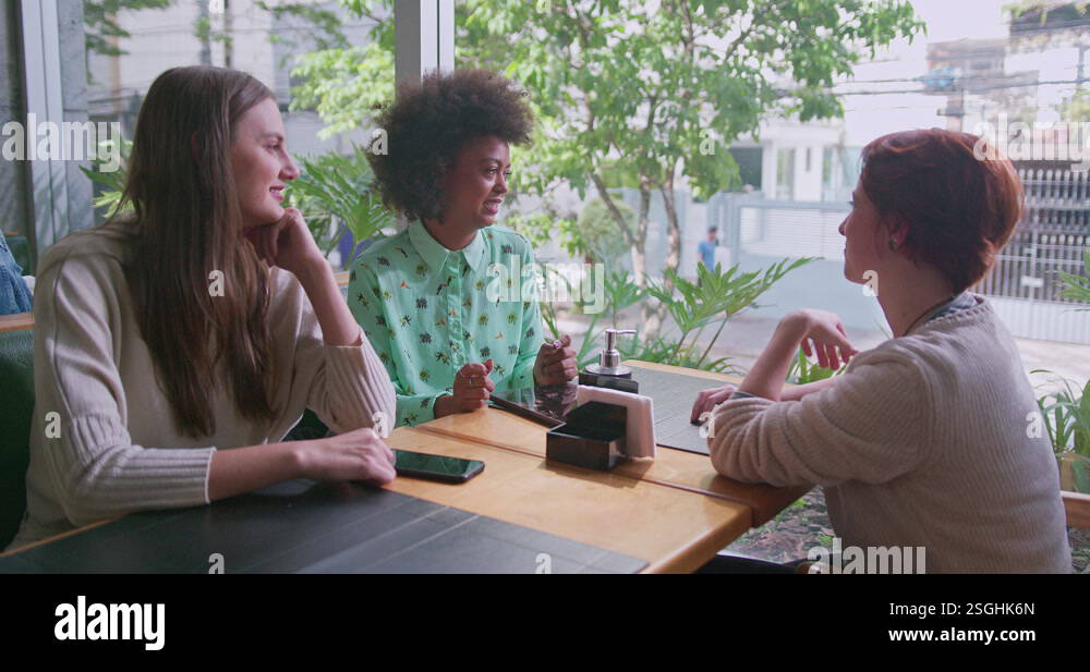Three happy diverse women sitting at coffee shop table by window ...
