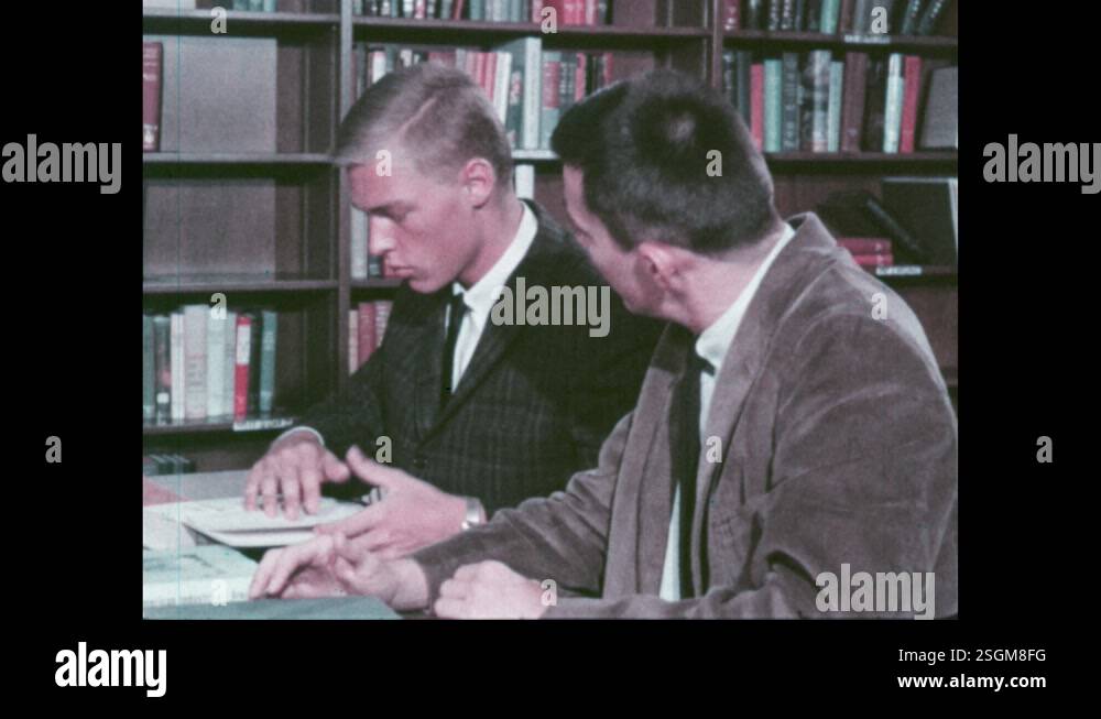 1960s:Finger points at title of book. Men sitting at table, look ...