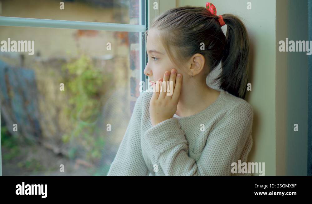 Scared sad girl refugee sits on windowsill and looks out window at ...