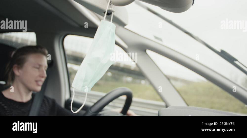 Female drives car face mask hang on rear mirror rainy day in close up ...