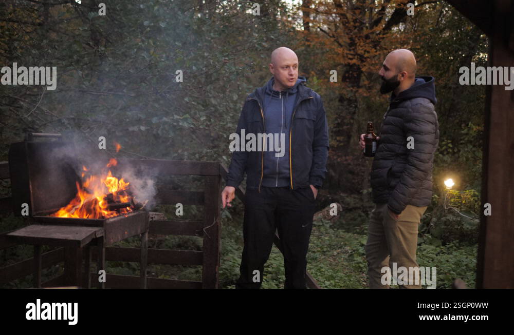 Two middle-aged adult men control the fire of logs in the barbecue talking and Stock Video ...