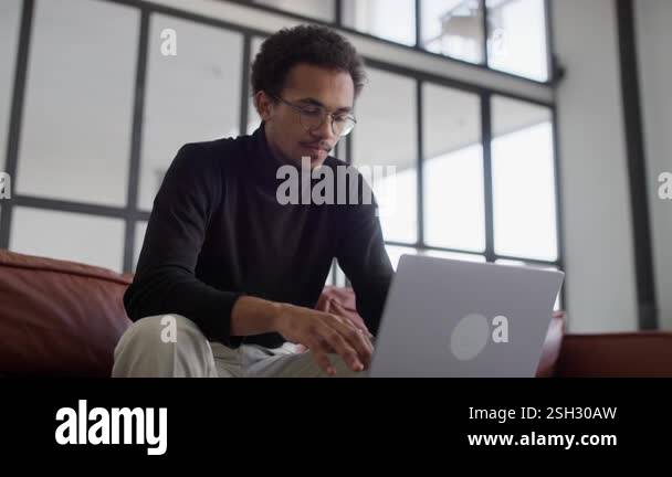 A young man sits on a leather couch in a modern office, looking tense ...