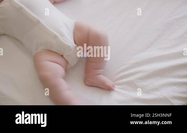 Chubby baby in white bodysuit, lying on a bed in a bright room, plays ...