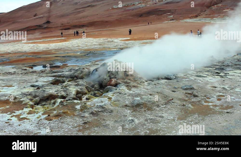A smoking volcanic geyser, with people and sandy brown mountains in the ...
