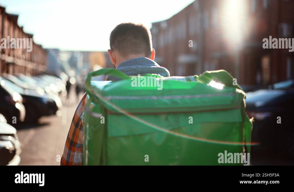 Rear view of male food delivery man walking down city street with ...