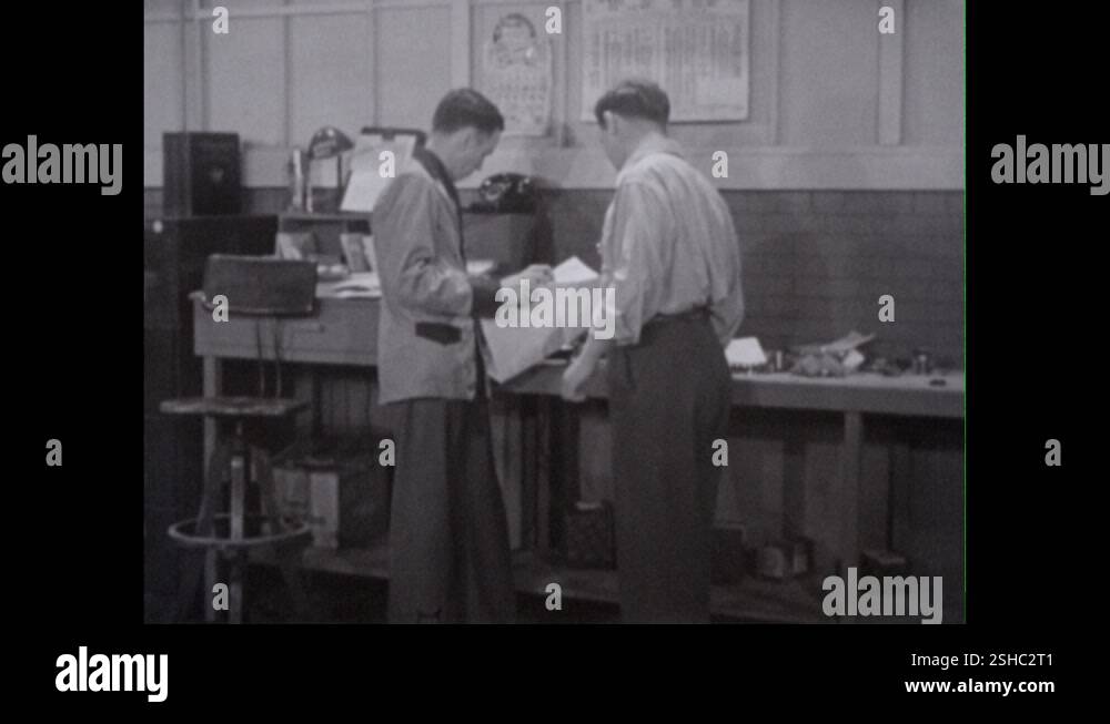 1940s: Foreman talks to worker at desk in factory. Man holds smoking ...