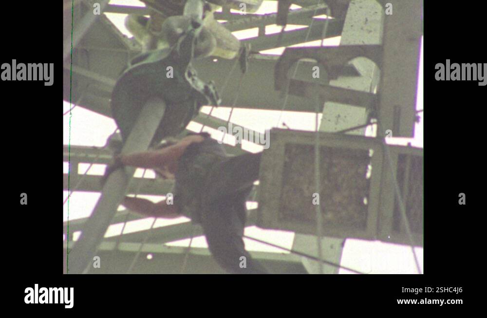 1970s: Oil well, tall platform, machinery rises on cable. Man hangs ...