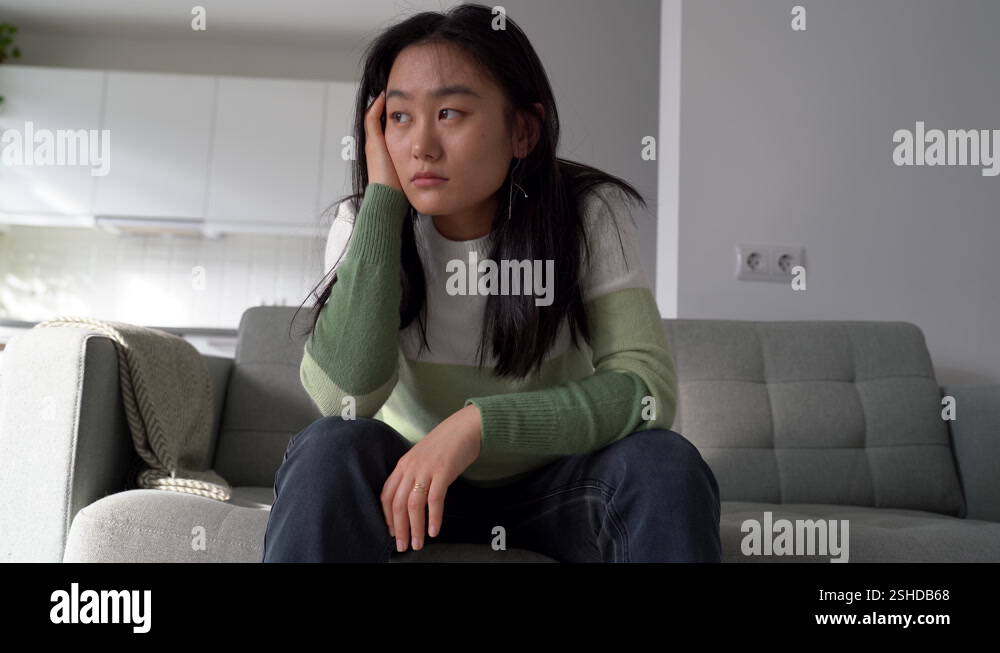 Worried Asian woman sits on sofa with concerned face expression ...