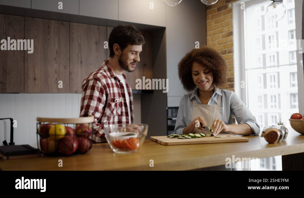Young man joining his girlfriend who's cooking in the kitchen, couple ...