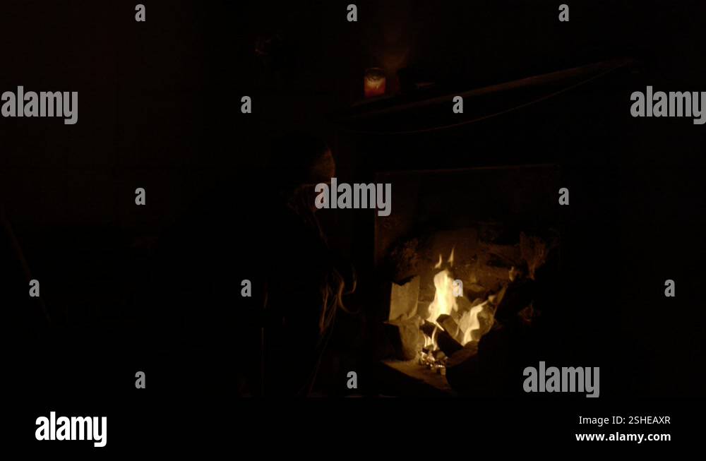 A man sits in a dark room warming himself by a log fire in an old ...