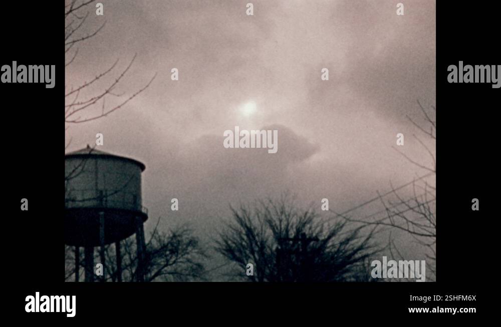 1970s: Sun shines behind clouds. Clouds move past water tower and trees ...