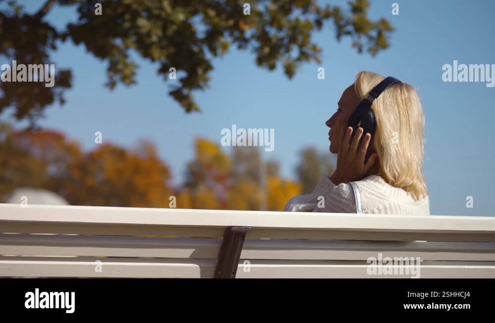 Back view of mature woman in headphones relax on bench in park ...