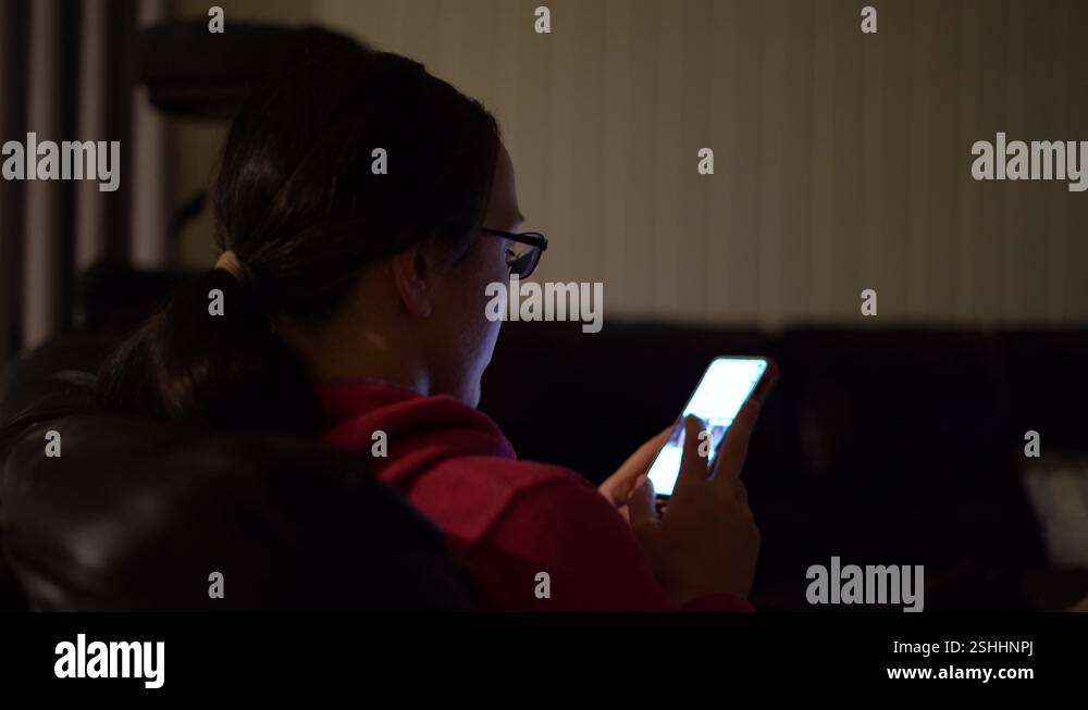 Mid aged woman sitting in sofa in poorly lit living room and scrolling ...