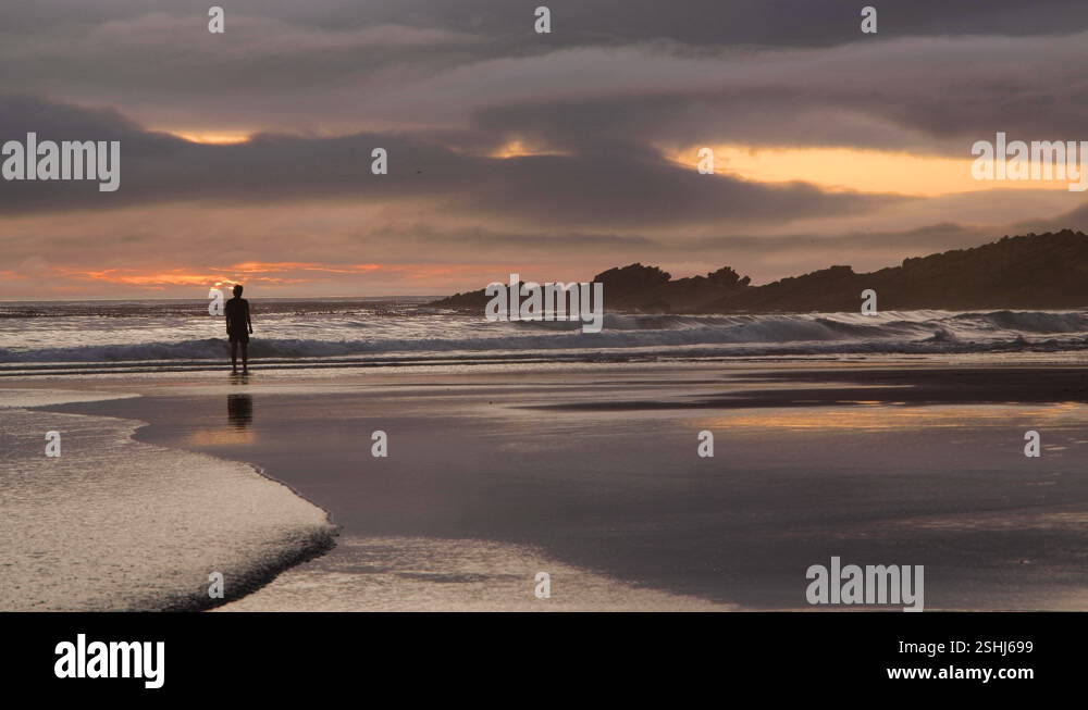 Silhouette of man standing in shallows on beach watching sunset; zoomed ...
