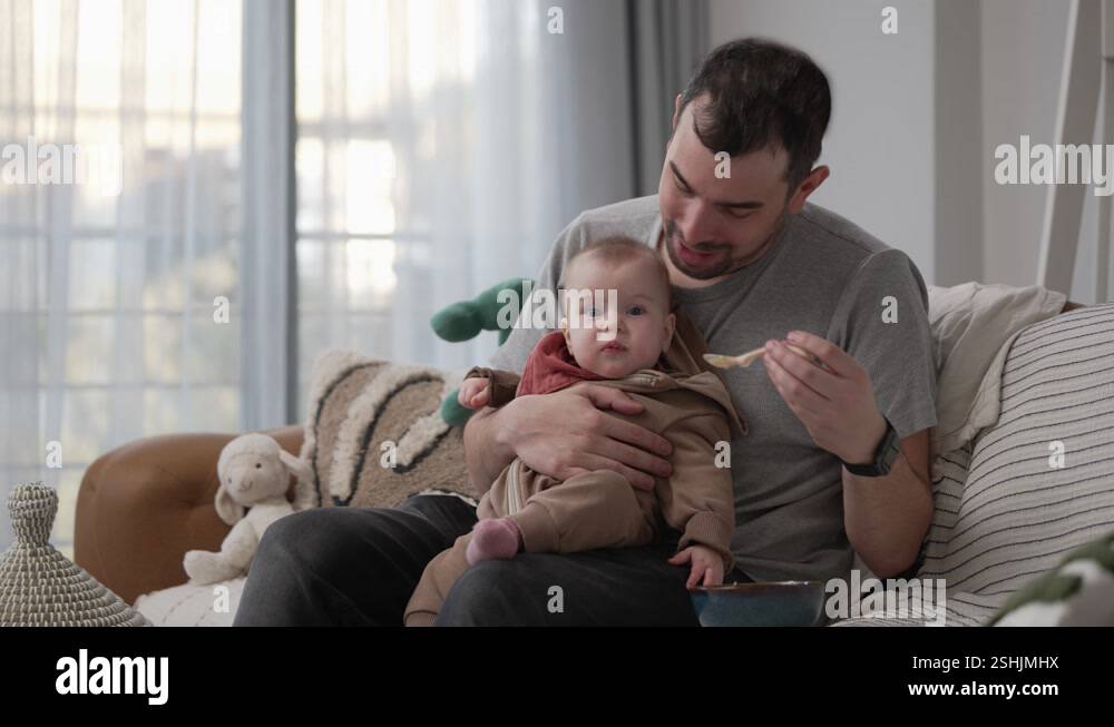 A Young Father Sitting on the Couth, Holds a Baby and Feeds him with ...