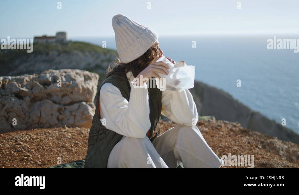 Girl hiker eating food at ocean cliff top. Serene travel breakfast at ...