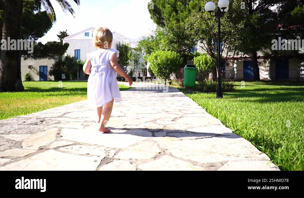 Little girl walks along a tiled path in a garden, balancing with her ...