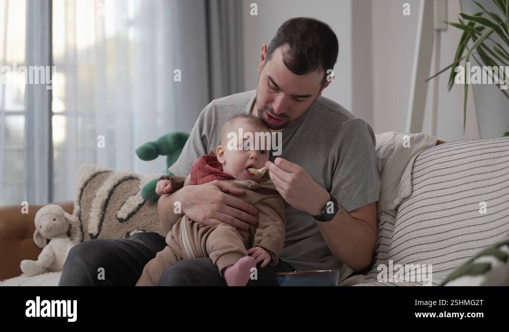 A Young Father Sitting on the Couth, Holds a Baby and Feeds him with ...