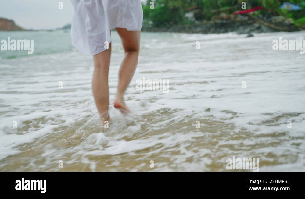 Slim female legs and feet walking along sea water waves on sandy beach ...
