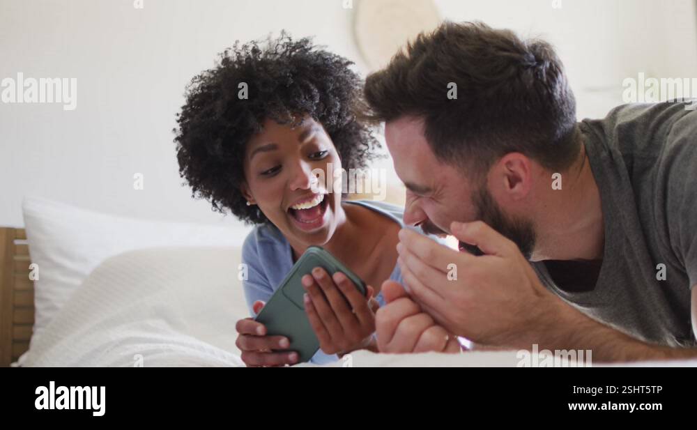 Video of happy diverse couple relaxing at home, lying on bed using ...