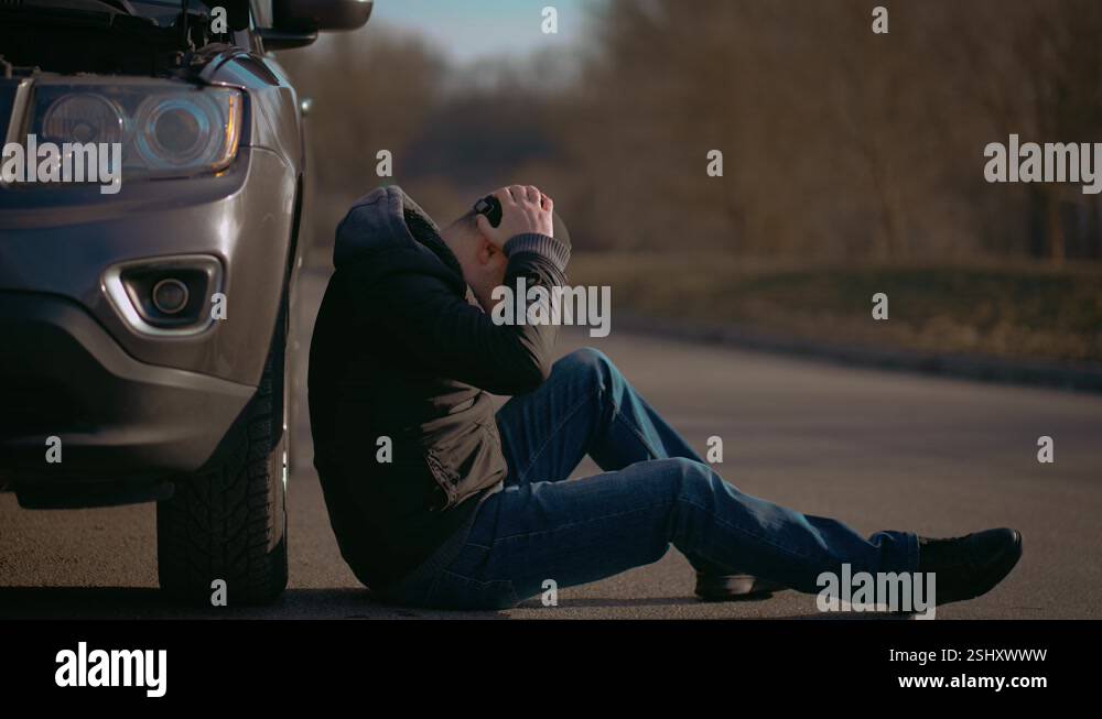 Sad depressed man sitting on road near broken car with opened hood ...