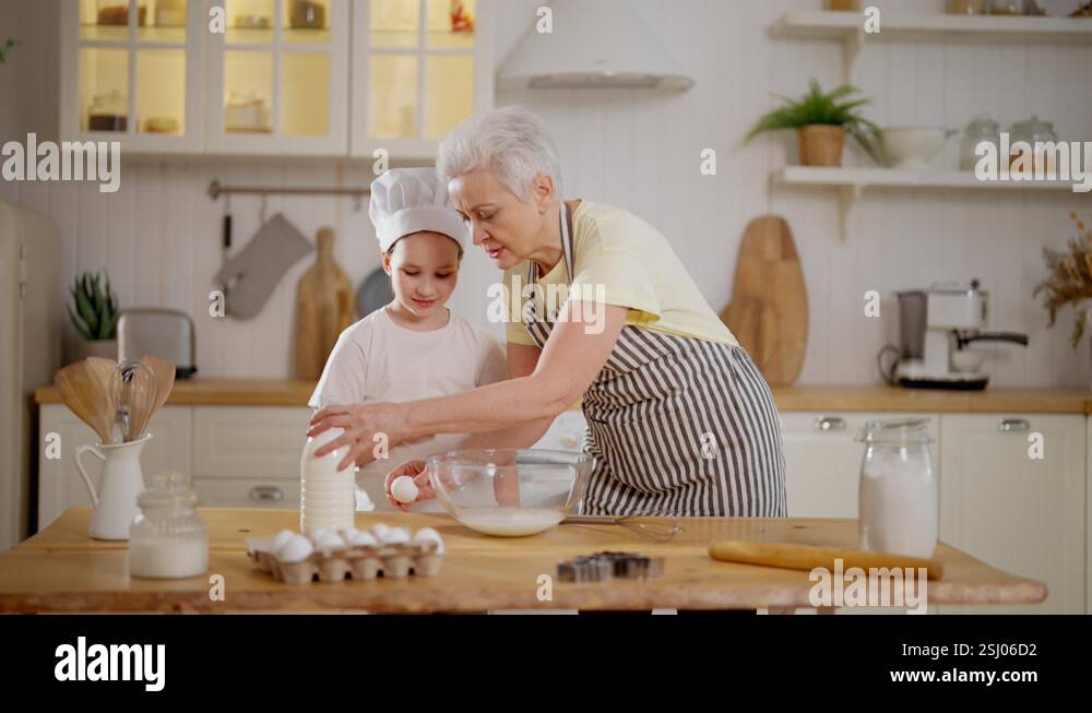 Granny and granddaughter cooking together adding eggs to milk in bowl ...