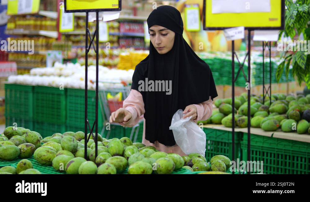 Young woman in hijab chooses ripest fruits in large supermarket. She ...