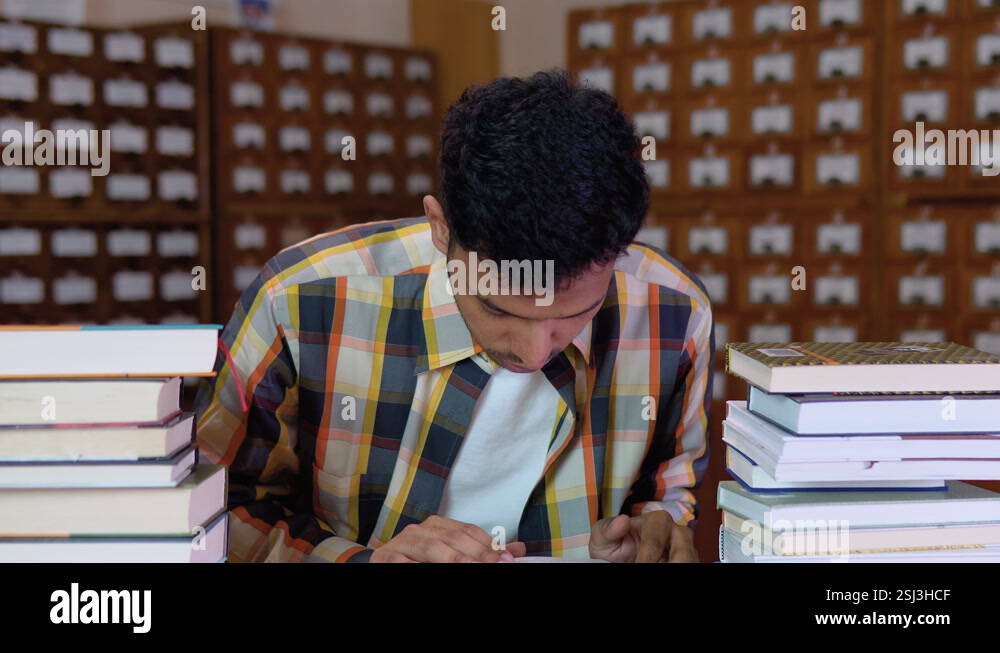 Tired indian student, in casual clothes, sits at a desk in a library ...