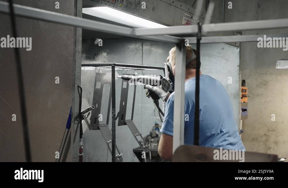 Blue collar worker spraying powder coating onto metal in an industrial ...