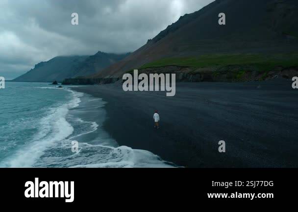 Lonely sad man in white coat and red hat walk on dark black volcanic ...