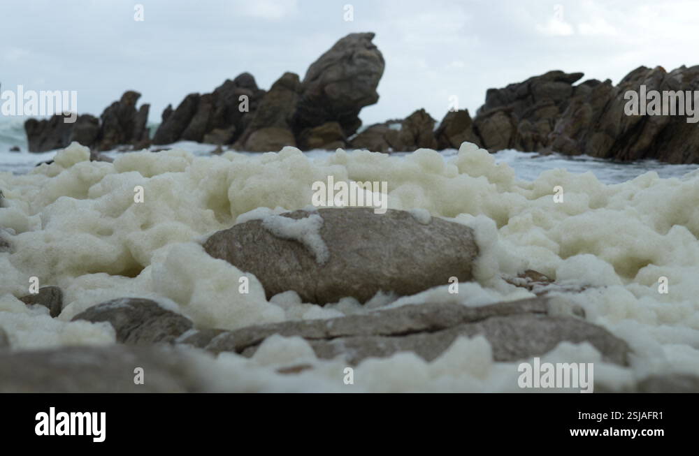 Sea foam blows in windy conditions on a rocky shore as rough sea waves ...