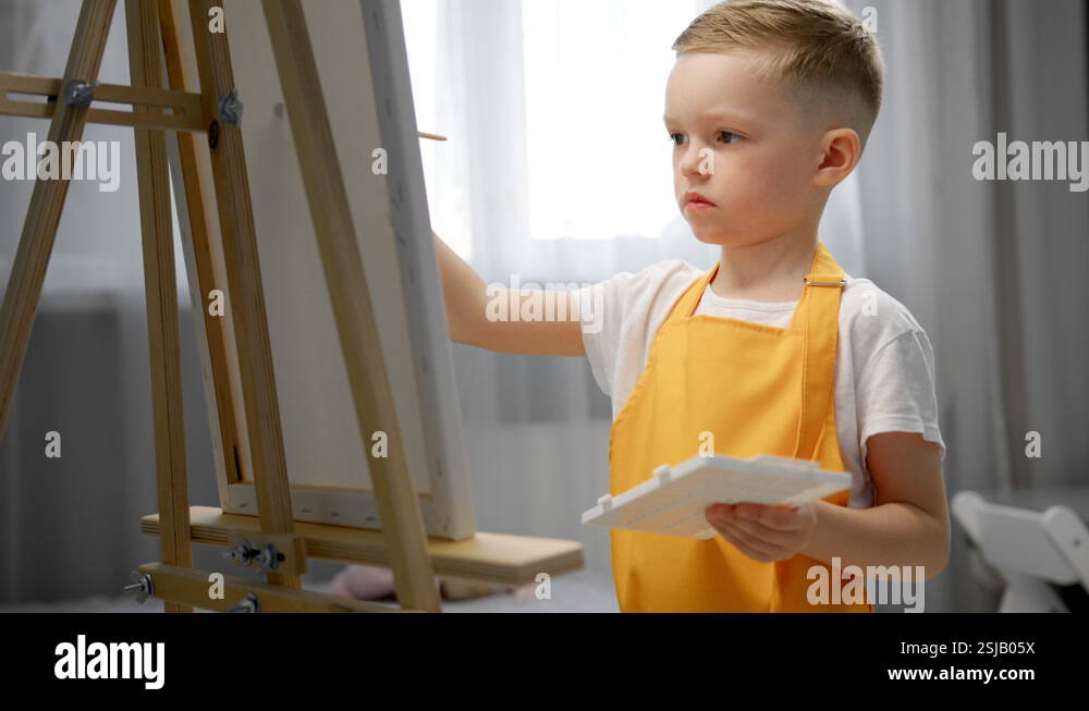 Talented blonde boy artist-innovator paints with brush on large canvas ...