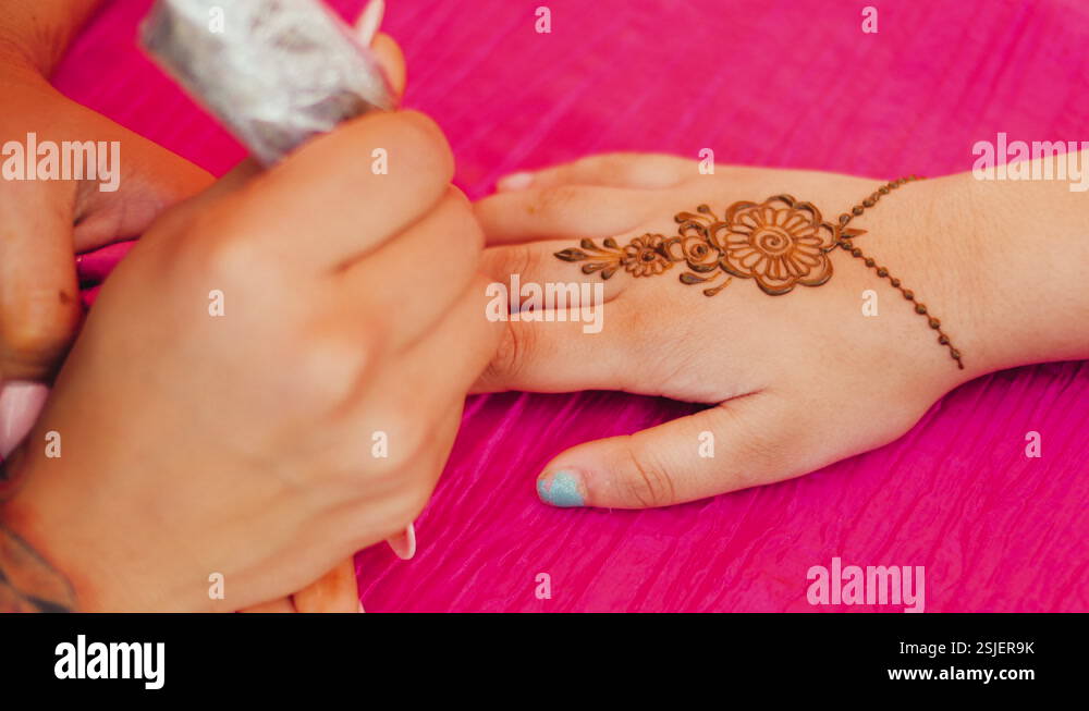 A close-up of a mehendi master draws henna on person's hand on a pink ...
