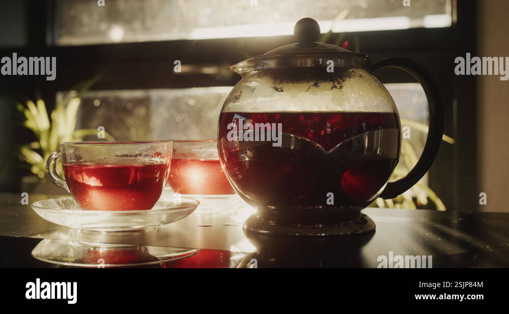 A glass teapot with tea and two cups stand by the window from which the ...