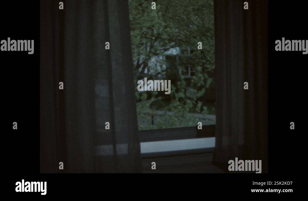 1960s: Looking out open window of house. Curtains inside house blow ...