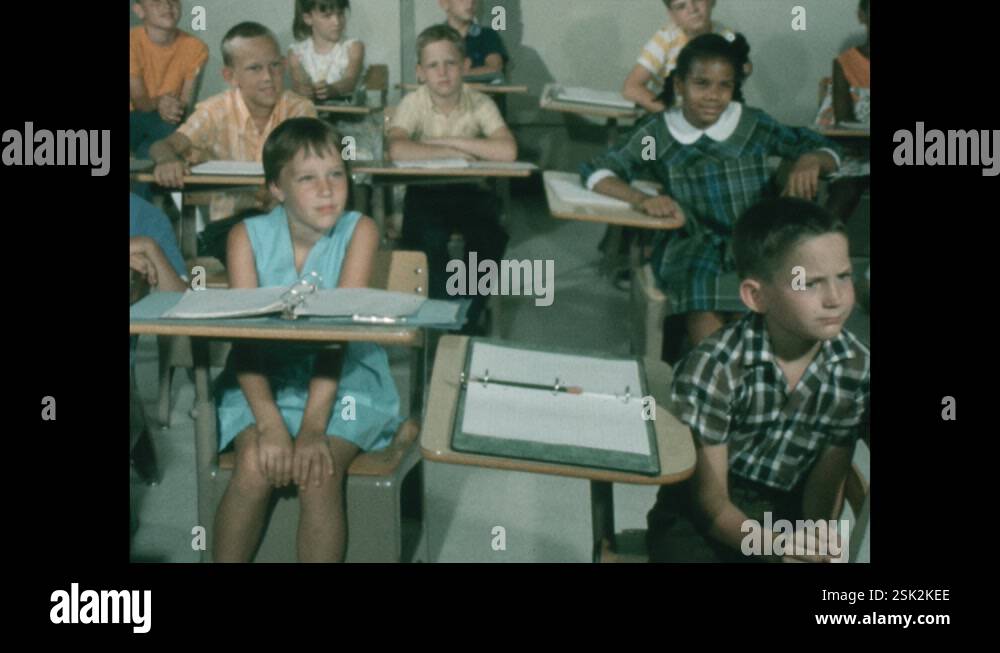 1960s: Students sit in classroom desks, listening. Young boy raises his ...