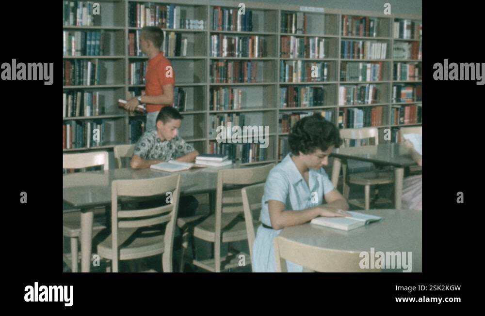 1960s: Students study at tables in library. Boy walks into library and ...