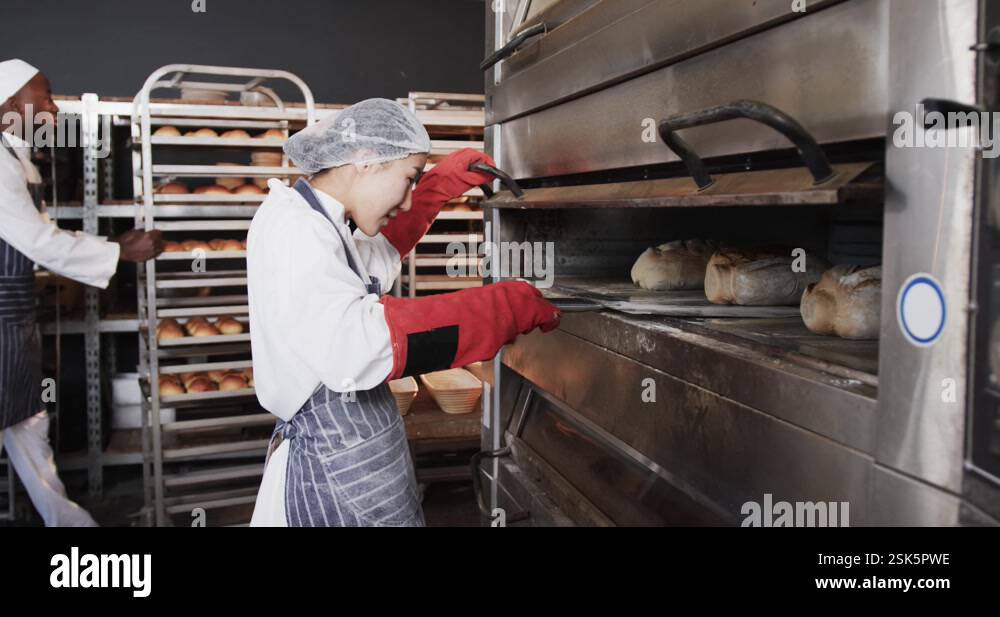 Asian female baker working in bakery kitchen, taking bread out of oven ...
