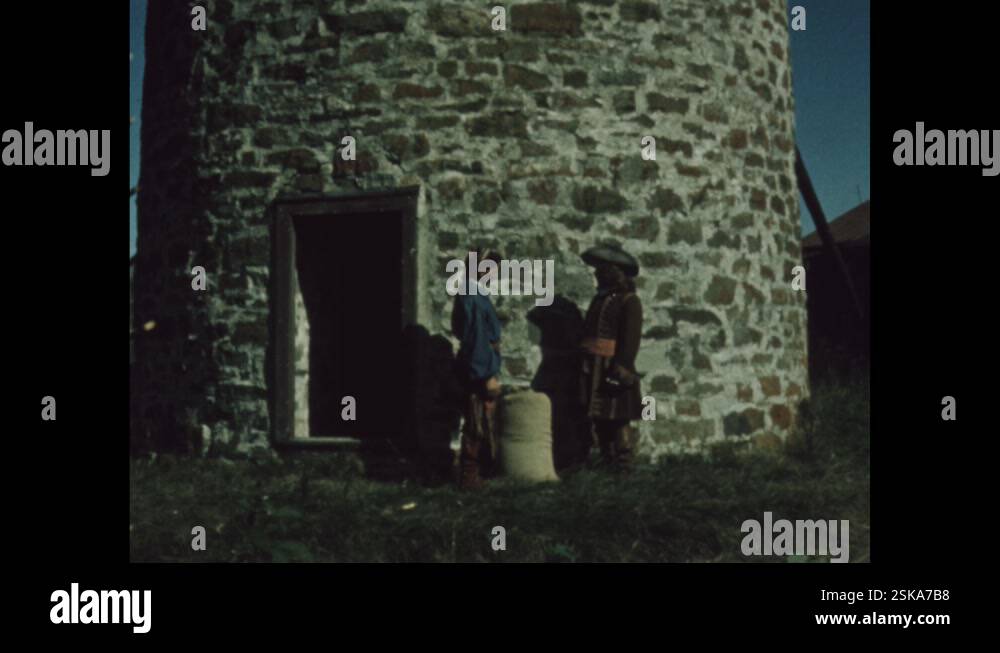 1960s: Two men stand outside windmill talking, one holds full sack over ...