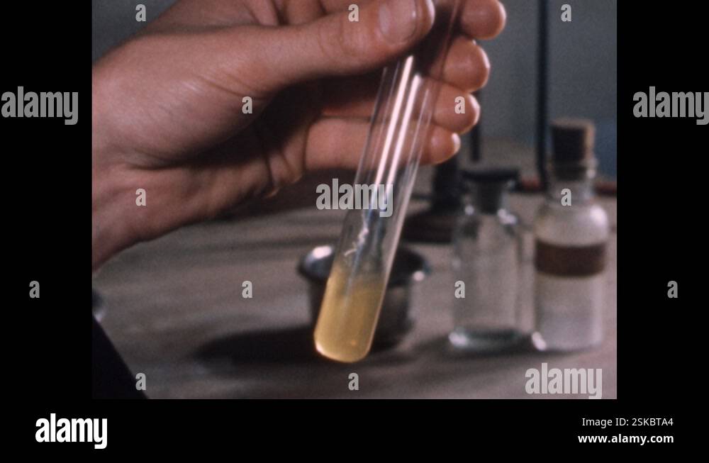 1960s: Scientist holds and shakes test tube of yellow liquid. Table ...