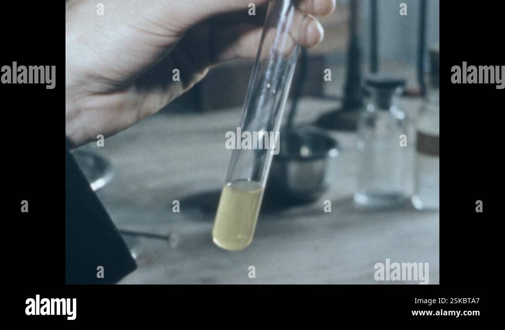 1960s: Scientist holds and shakes test tube of yellow liquid. Table ...