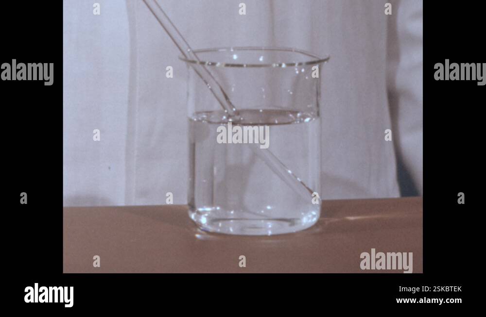 1960s: Glass rod sits in beaker of clear liquid. Hands add green food ...