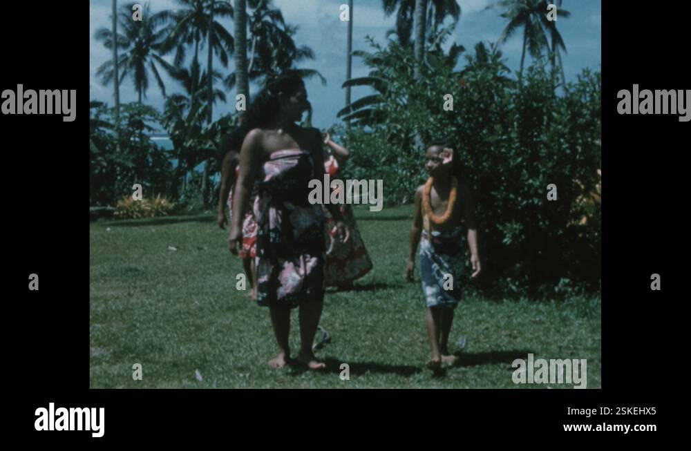 1960s: Women and children in sarongs with flowers in their hair walk ...