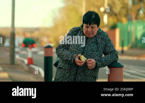 Woman leaning on bollard eating fruit near suburban street. Old female ...