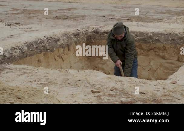 A man wearing a hat and jacket is digging a deep hole in the sandy ...