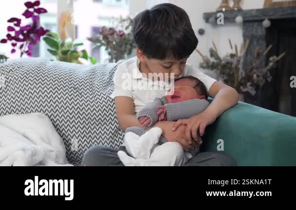 Young boy kisses his crying newborn sibling's head, both nestled in a ...