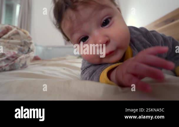 Baby lying on stomach on bed, looking directly at camera with curious and engaged expression ...