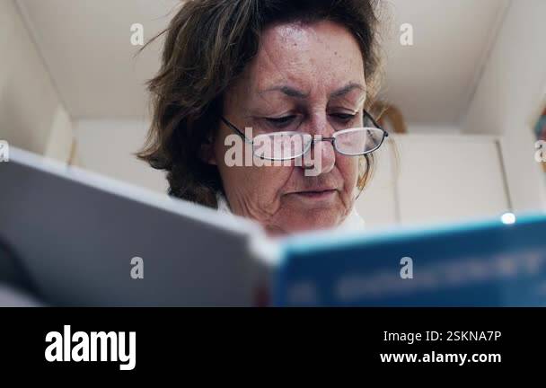 Elderly woman wearing glasses is deeply focused on reading a book ...