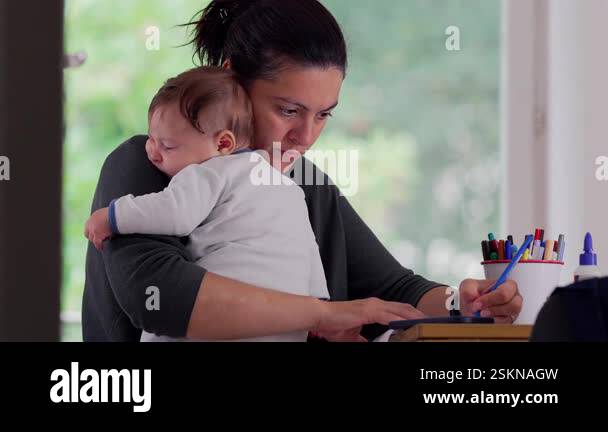 Mother holding her baby while writing at a desk, focused on completing ...